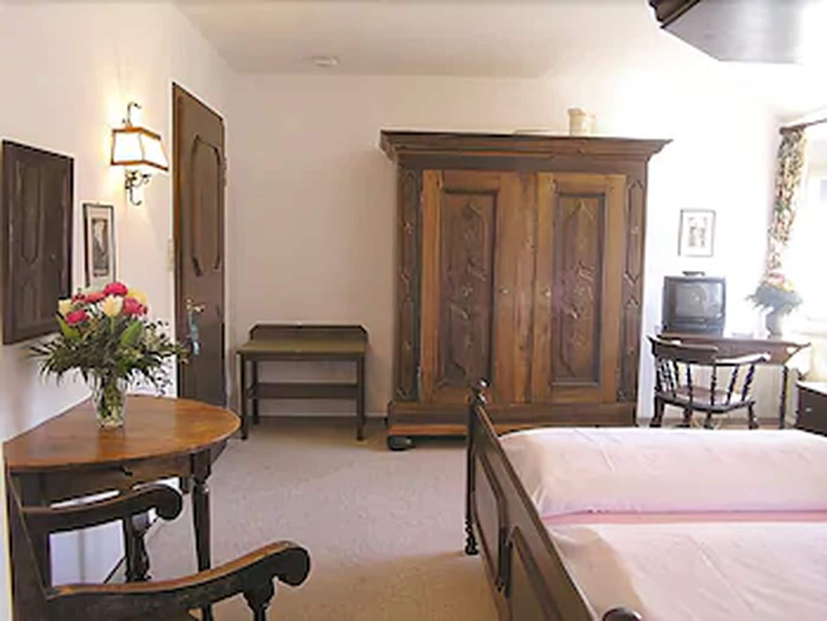 Um quarto com uma cama, um armário de madeira, uma mesinha com uma luminária e uma televisão sobre um criado-mudo.