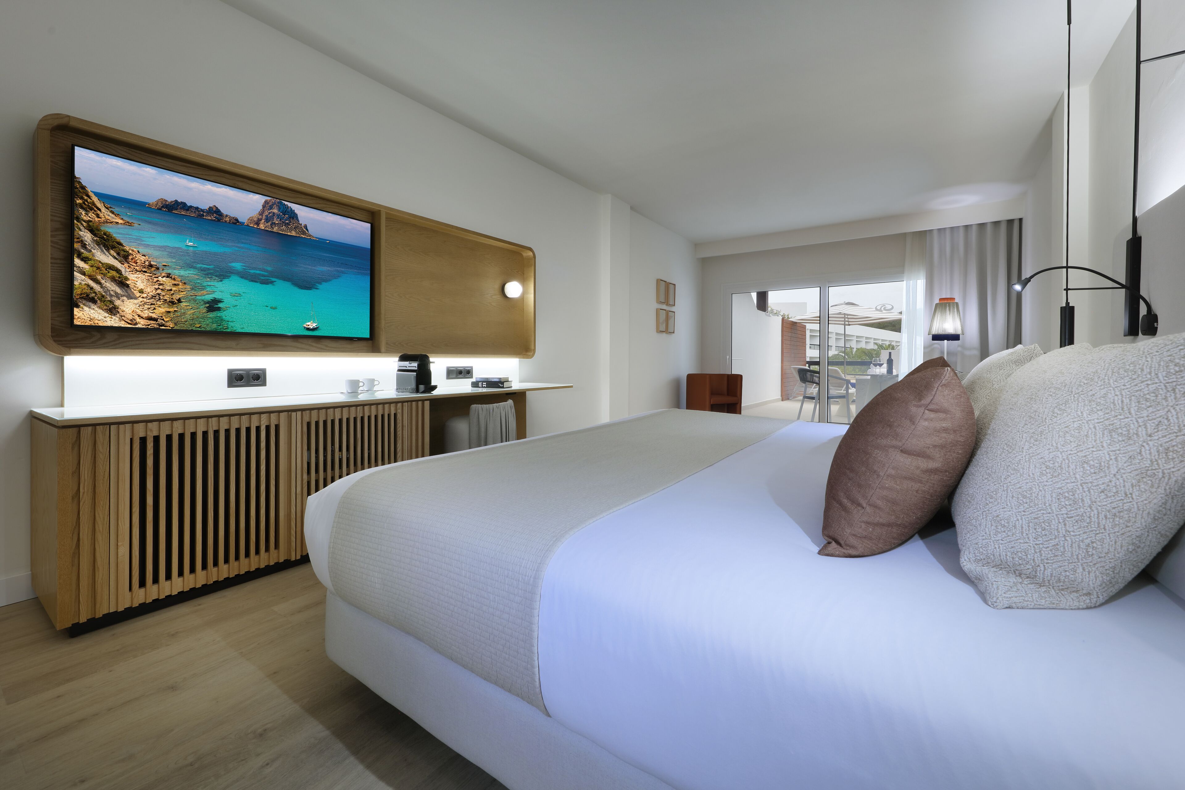 Quarto de hotel moderno com uma cama grande, uma TV presa a um console de madeira, e vista para o mar pela janela.