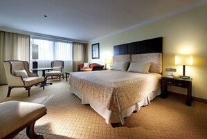 Quarto de hotel com uma cama grande, duas poltronas, uma mesa pequena e uma escrivaninha com abajur.