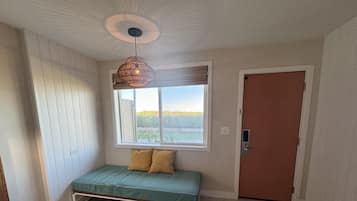 Um quarto pequeno com uma cama de solteiro, janela com persianas, porta e luminária no teto.