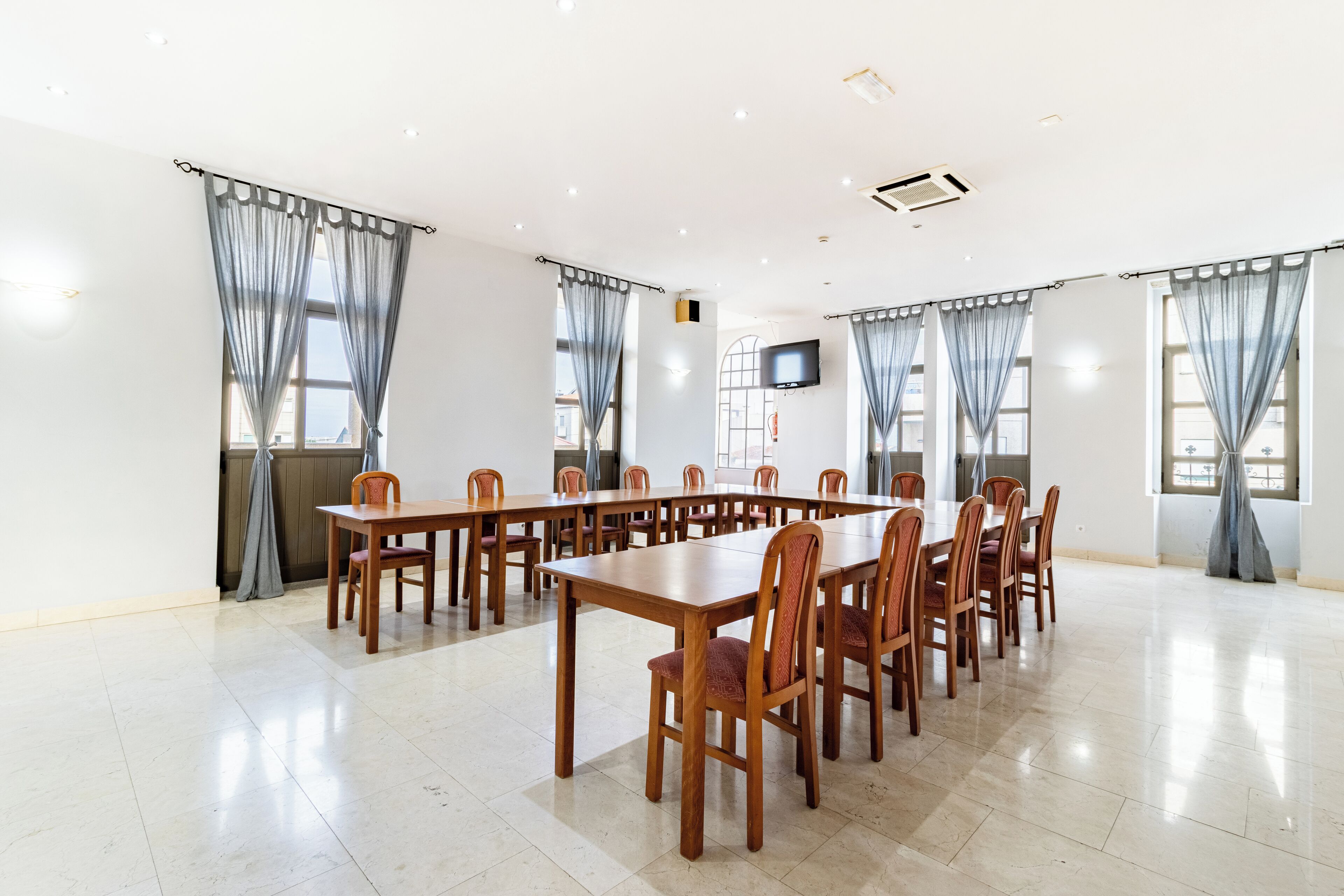 Uma sala de conferências com uma mesa comprida e cadeiras, janelas amplas com cortinas e uma TV de tela plana fixada na parede.