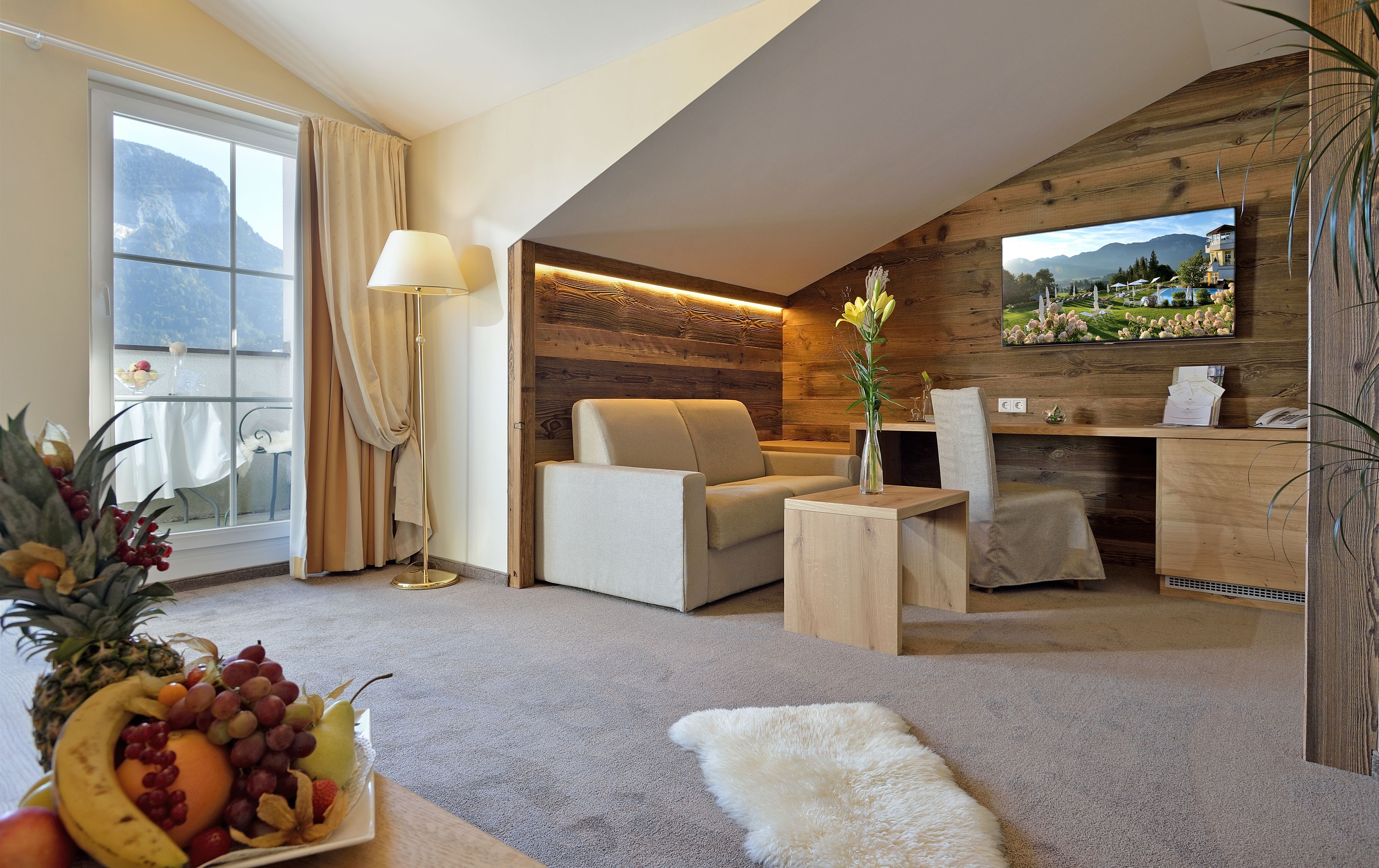 Uma sala de estar moderna com sofá, poltronas, mesa de centro e televisão montada numa parede de madeira. Uma janela grande com cortinas é visível, e uma fruteira está no chão.