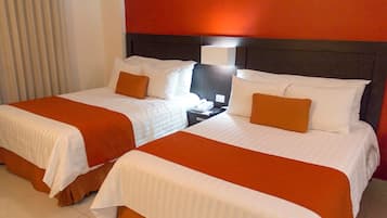 Quarto de hotel com duas camas, cada uma com roupa de cama branca e detalhes em laranja, uma mesa de cabeceira com abajur e uma parede em tom de vermelho.