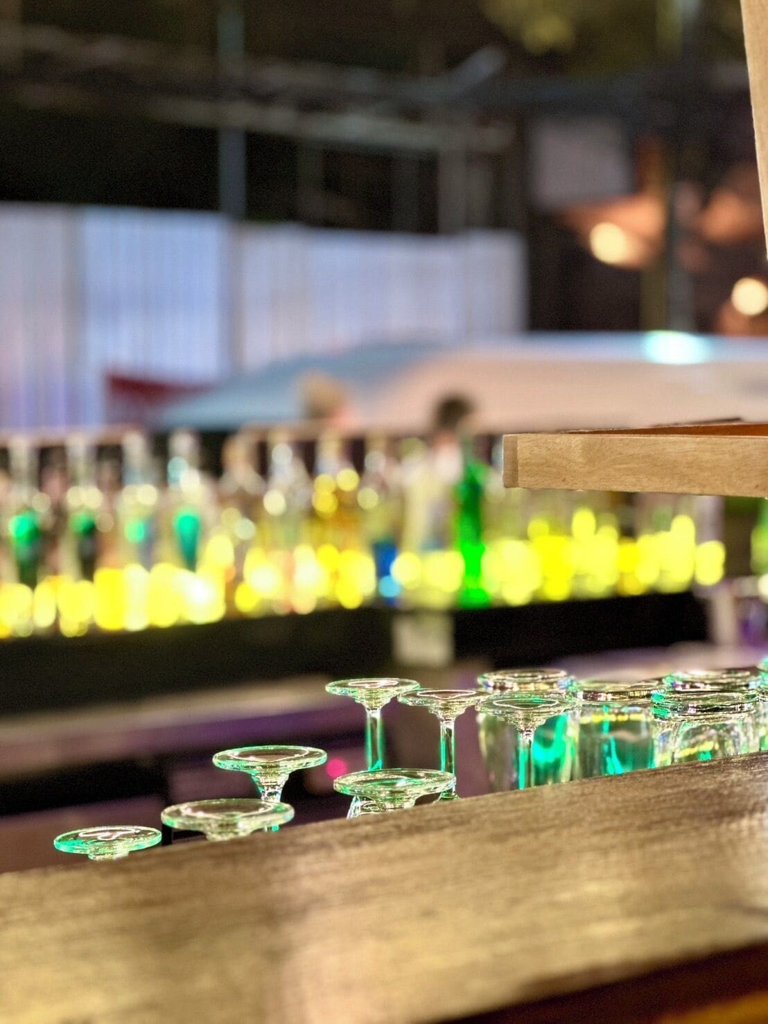 Um bar com vários copos e garrafas, prateleiras de madeira e um fundo desfocado com luzes.