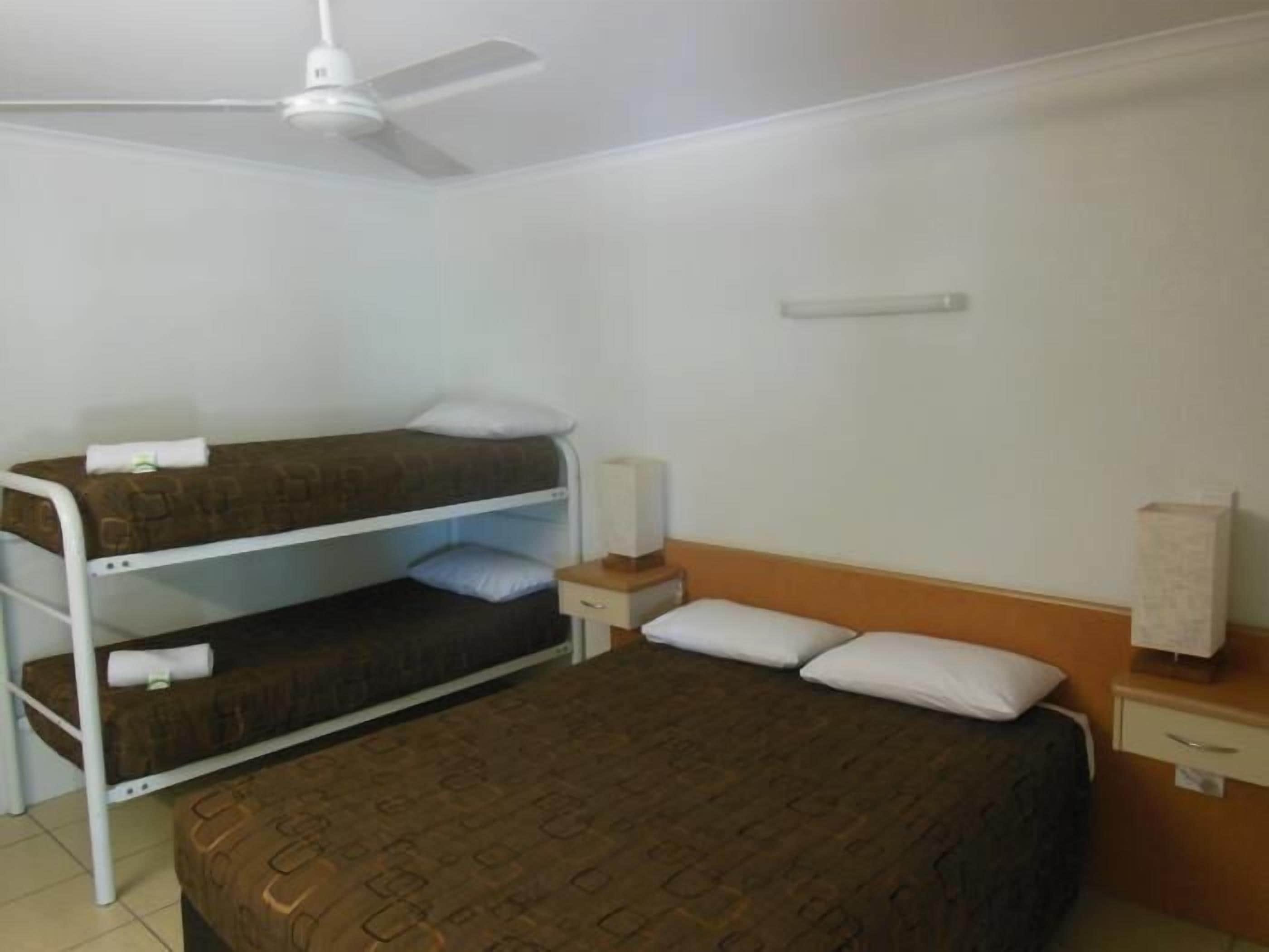 Quarto individual com cama de solteiro, beliche, ventoinha no teto, mesa de cabeceira com abajur e um pequeno armário.