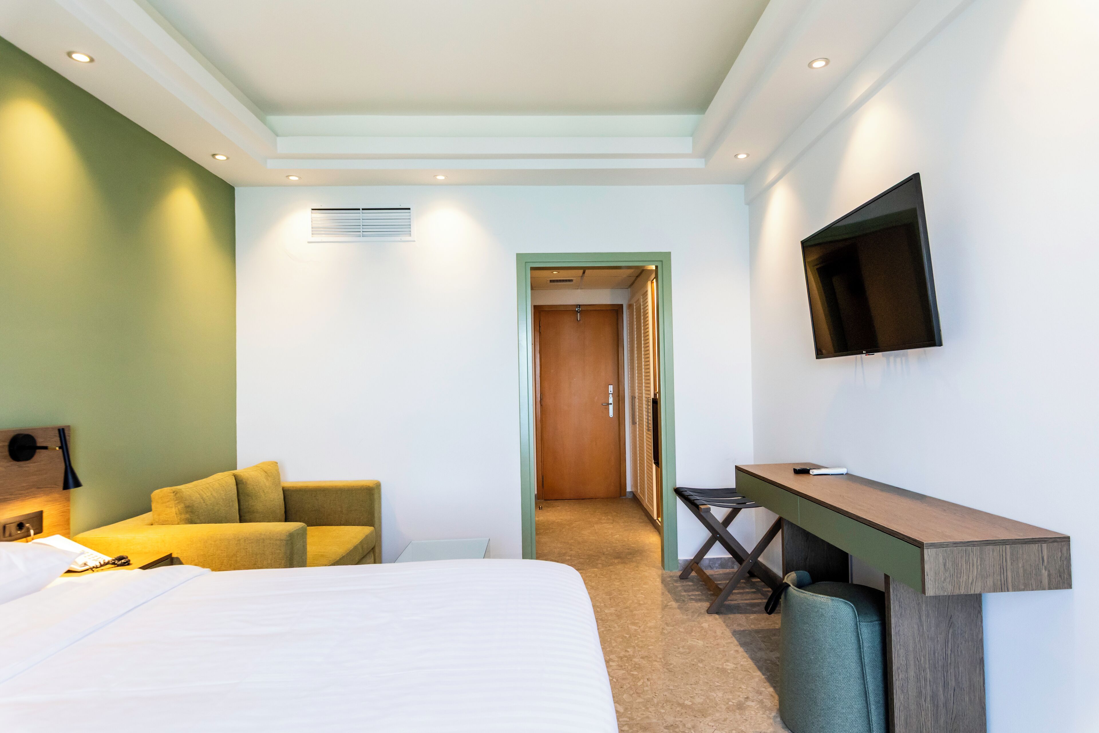 Quarto de hotel com cama, escrivaninha, cadeira, televisão e porta que dá para outro cômodo.