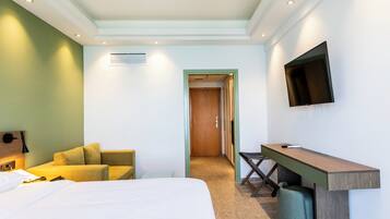 Quarto de hotel com cama, escrivaninha, cadeira, televisão e porta que dá para outro cômodo.
