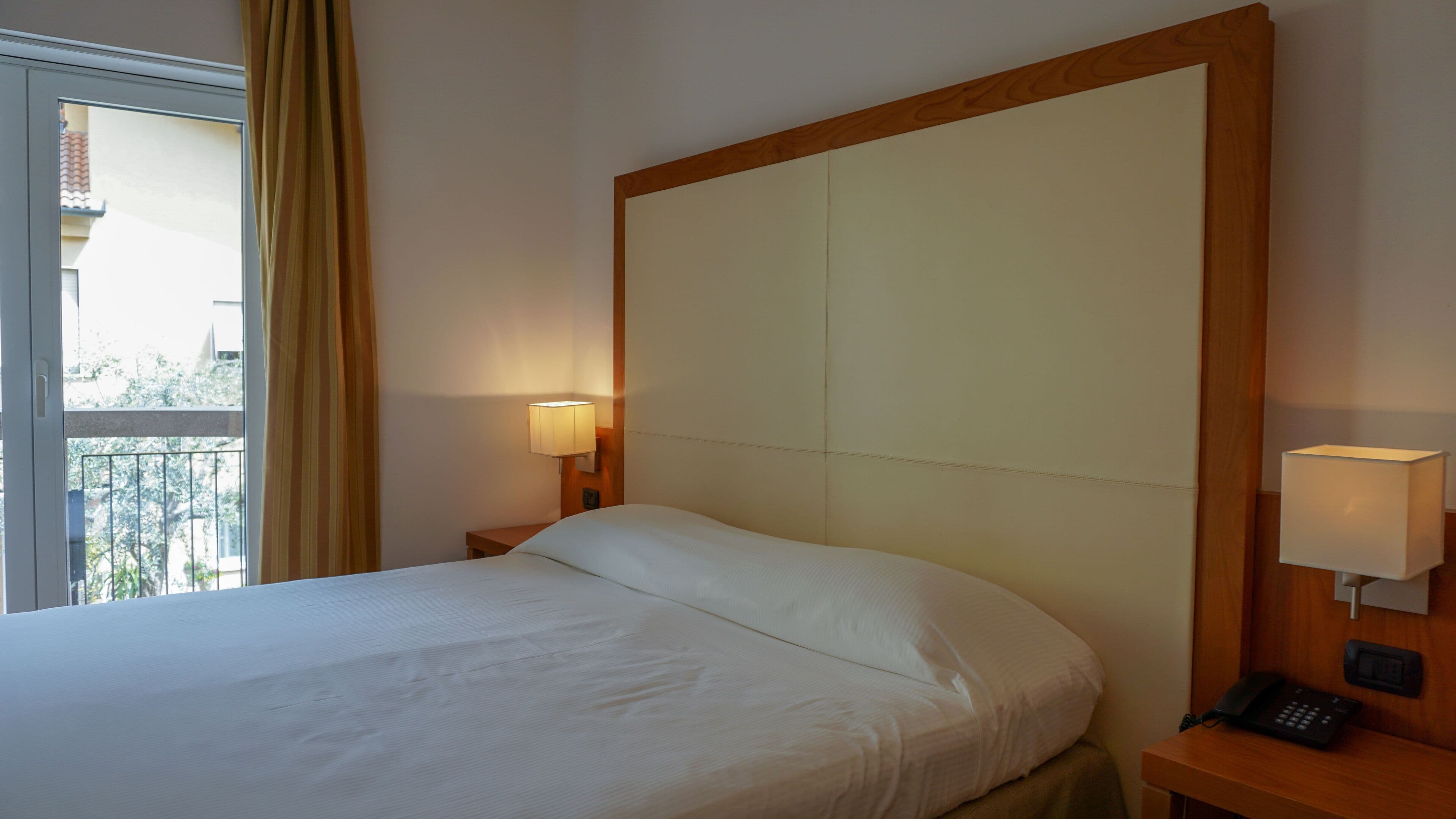 Quarto de hotel com uma cama grande, cabeceira de madeira, mesa de cabeceira com abajur e telefone.