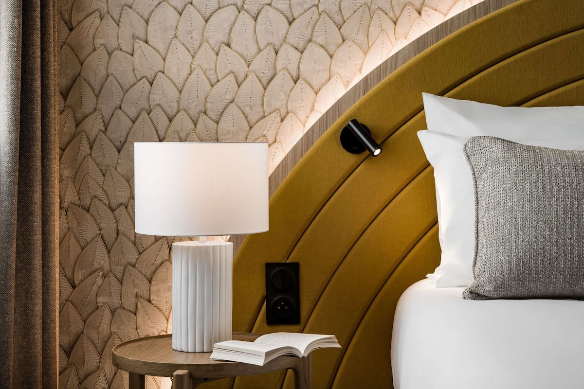Quarto de hotel com papel de parede estampado, mesa de cabeceira redonda com abajur, um livro e uma cama com cabeceira, além de painel de controle fixado na parede.