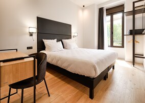 Quarto de hotel moderno com uma cama grande, uma escrivaninha e uma cadeira. Há uma janela com cortinas e vista para o verde lá fora.