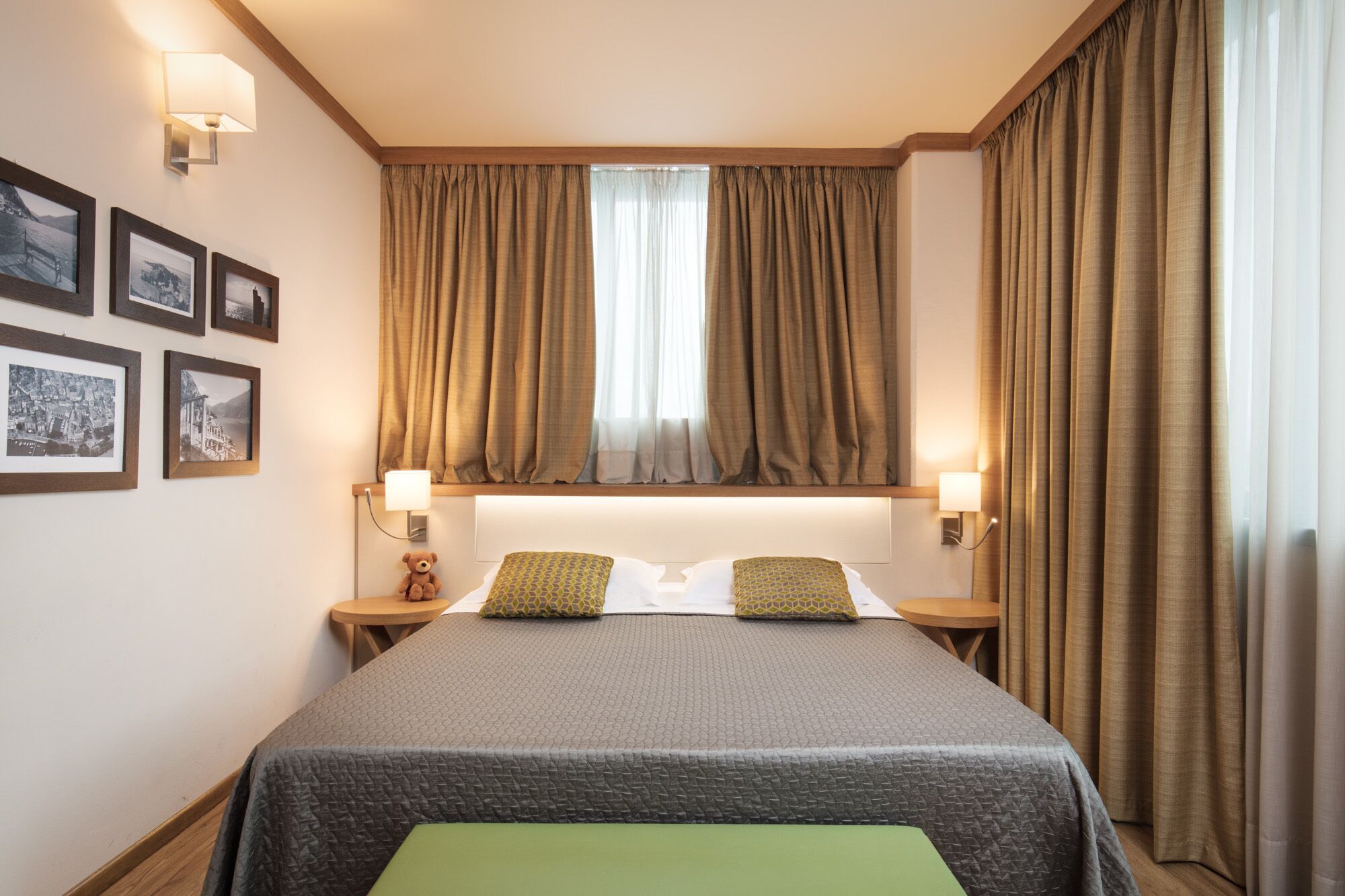 Quarto de hotel com uma cama grande, duas mesas de cabeceira, uma luminária na parede e uma janela com cortinas.