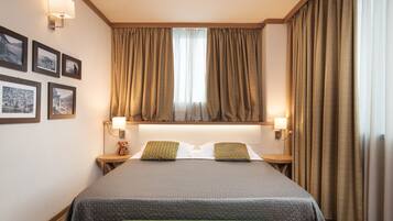 Quarto de hotel com uma cama grande, duas mesas de cabeceira, uma luminária na parede e uma janela com cortinas.