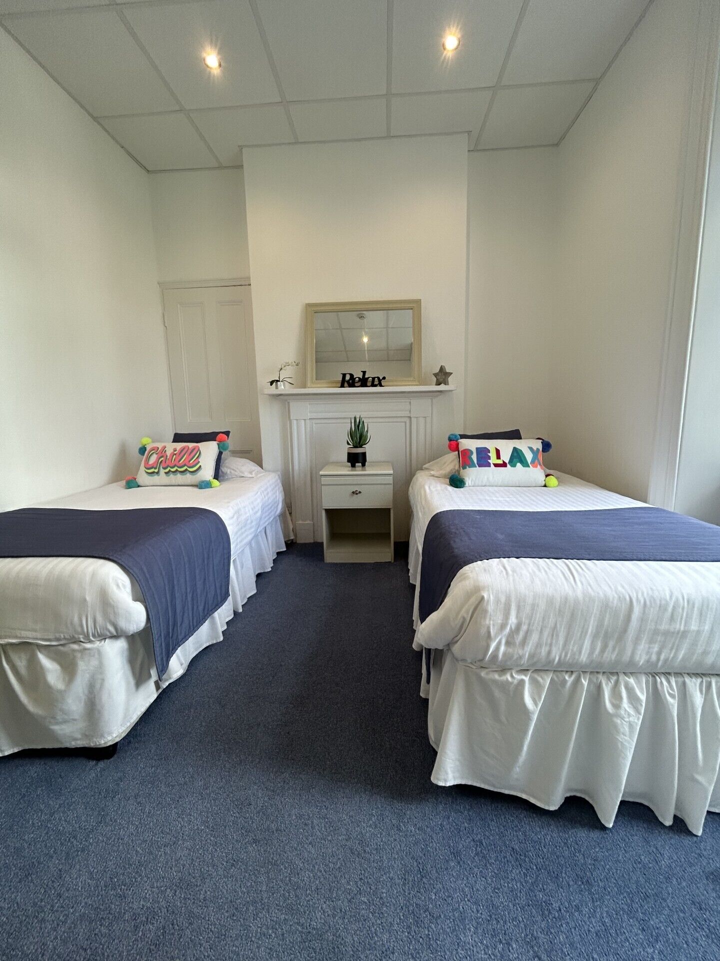 Quarto de hotel com duas camas de solteiro, uma pequena mesa de cabeceira com uma planta, um espelho sobre a lareira e uma porta à esquerda.