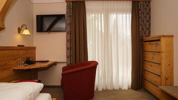 Quarto de hotel com cama, poltrona vermelha, penteadeira de madeira, escrivaninha com luminária e TV fixada na parede.