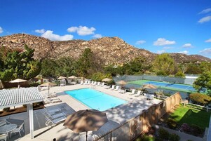 Piscina com espreguiçadeiras, quadra de tênis e montanhas ao fundo.