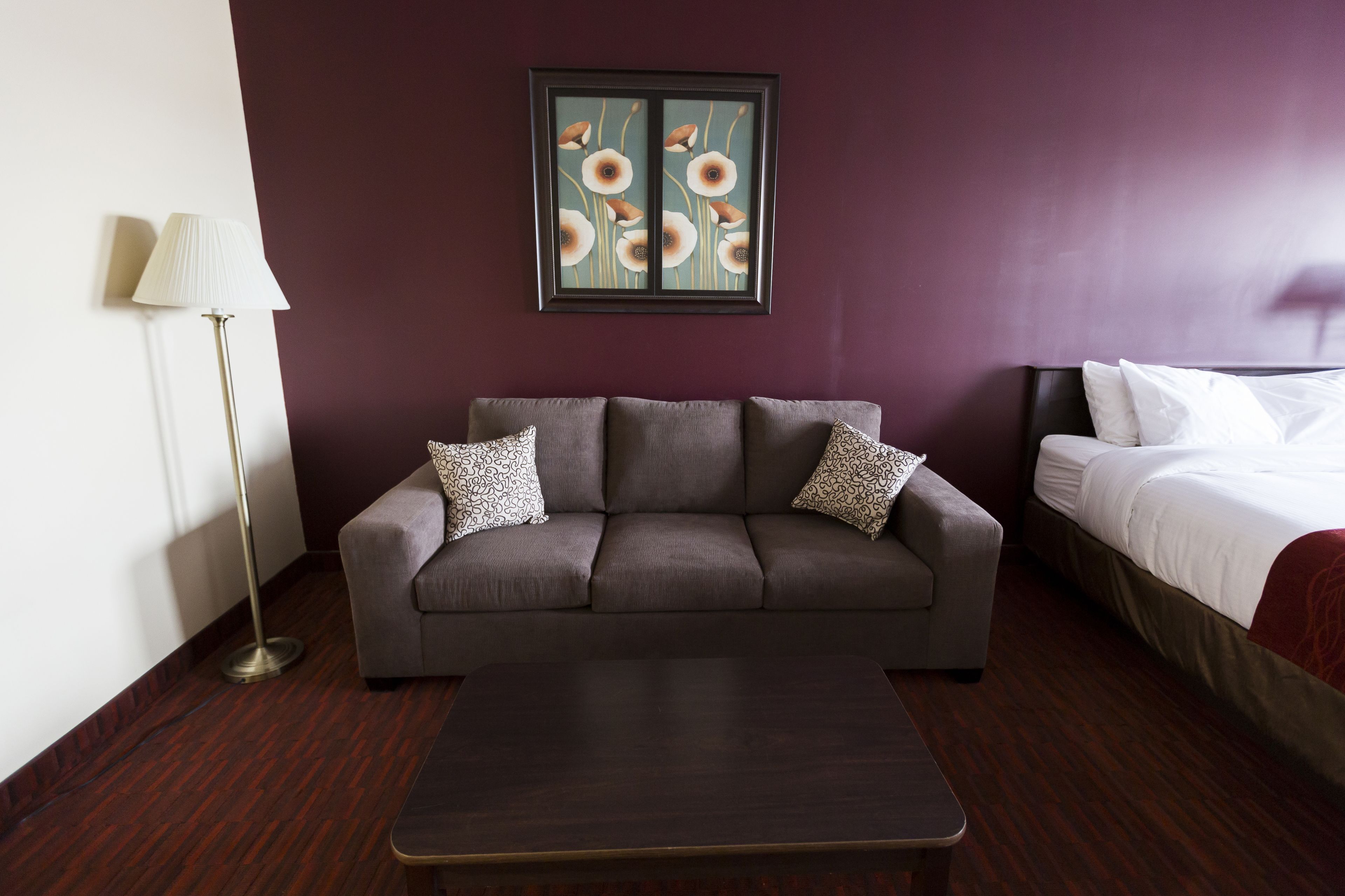 Um quarto com sofá cinzento, uma mesa pequena, uma cama com roupa de cama branca, um abajur de chão e uma obra de arte emoldurada na parede.