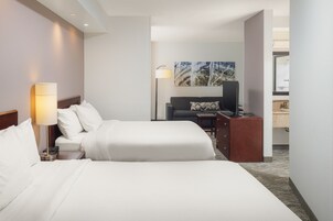 Quarto de hotel com duas camas, uma mesa, uma cadeira, um abajur, um quadro e um banheiro visível por uma porta aberta.