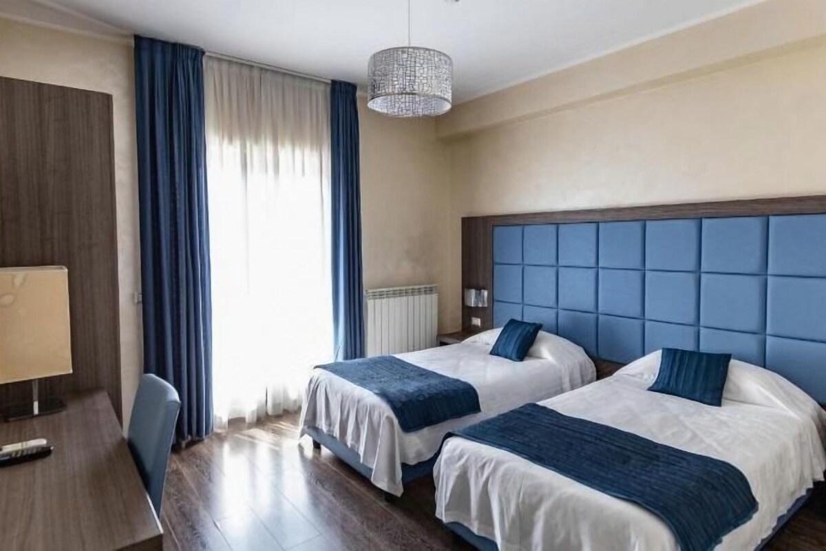 Quarto de hotel com duas camas, uma escrivaninha, uma cadeira, uma televisão e uma janela com cortinas.