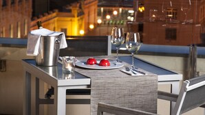 Uma mesa de jantar com uma garrafa, copos e uma vela numa varanda com vista para uma paisagem urbana ao entardecer.