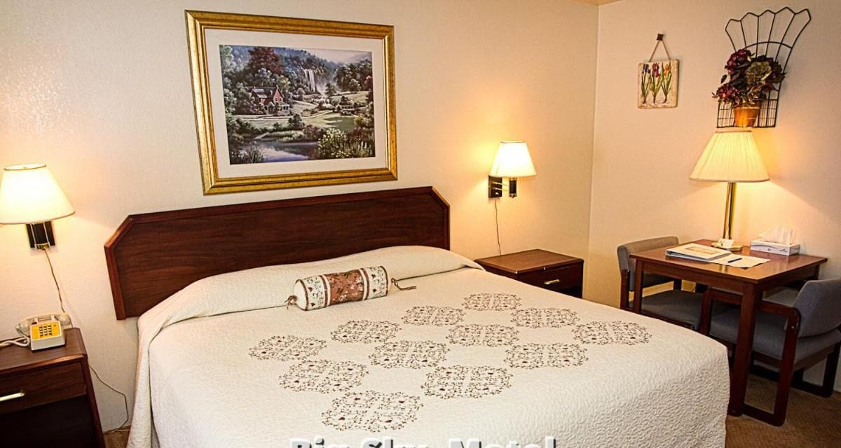 Quarto de hotel com uma cama grande, duas mesas de cabeceira, uma escrivaninha com cadeira e uma luminária na parede.