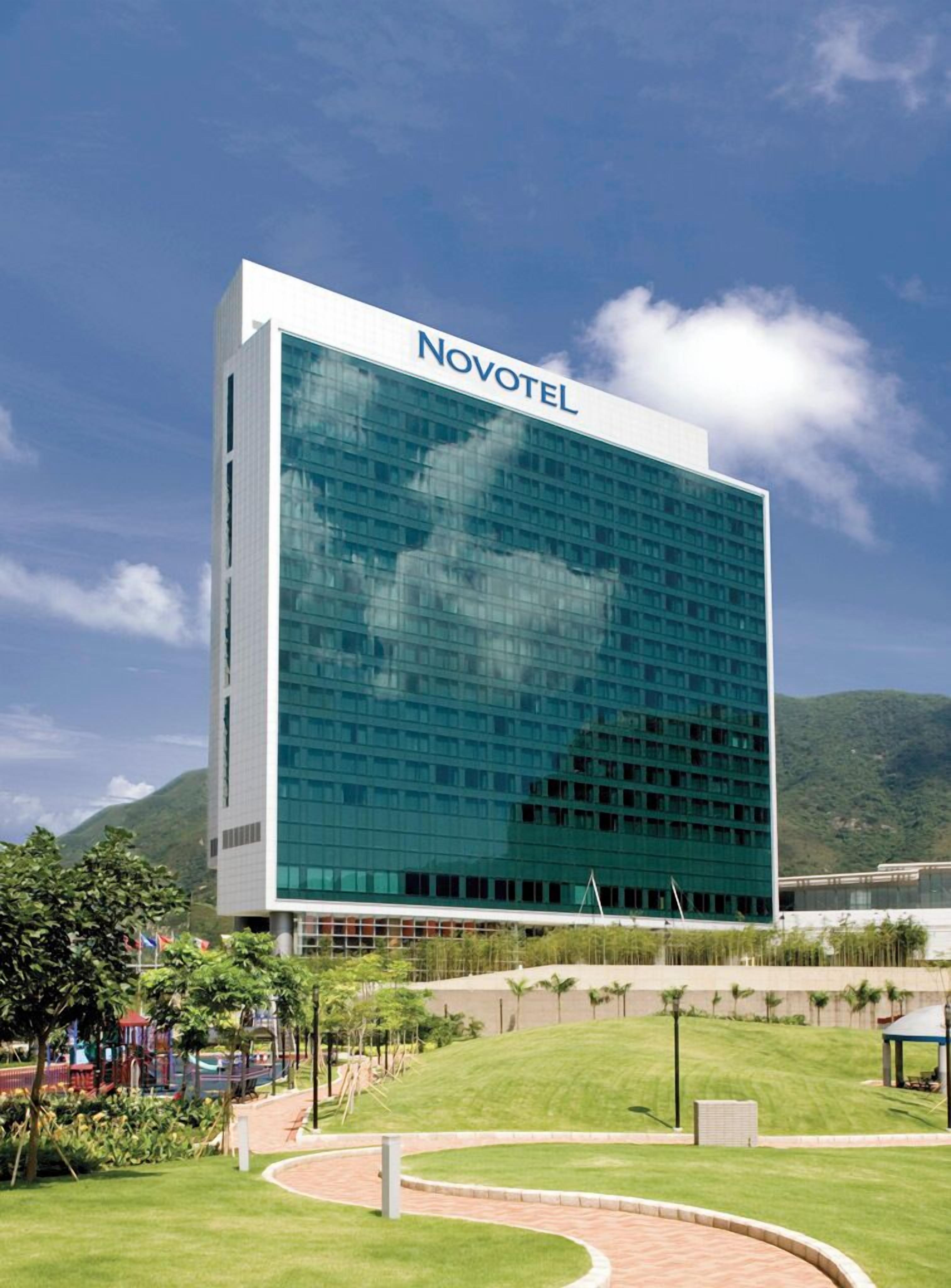 Um hotel Novotel com fachada de vidro moderna, rodeado por um jardim paisagístico e um parquinho.