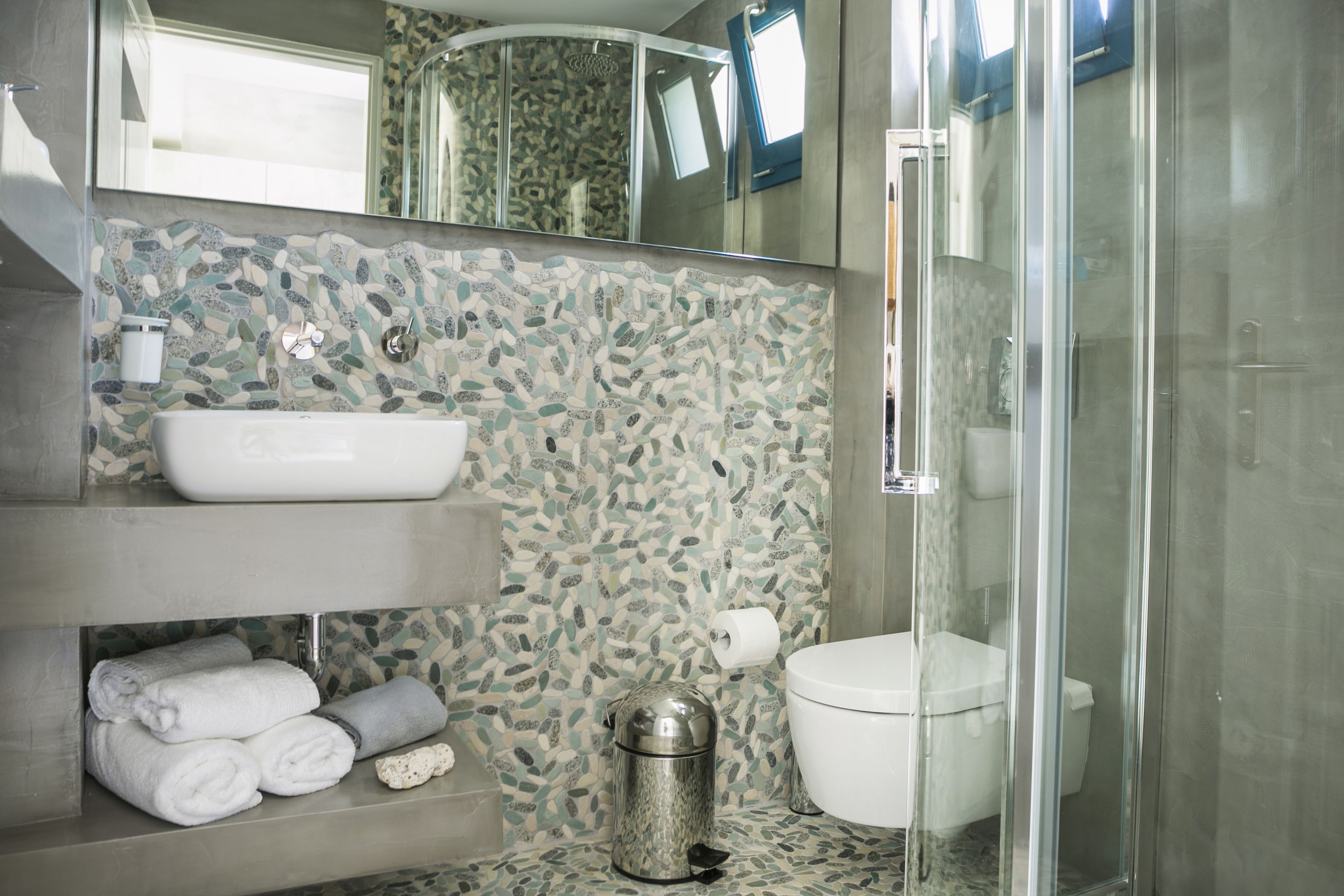 Casa de banho com painel de azulejos mosaicos, lavatório branco, sanita e cabine de chuveiro com portas de vidro.