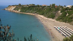 Uma praia com pessoas nadando e tomando sol, uma fileira de guarda-sóis e casas numa encosta.