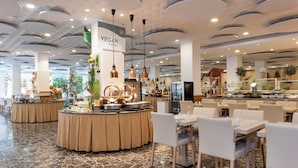 Uma área de refeições moderna, com mesas e cadeiras redondas, um balcão de buffet e uma placa que indica uma seção vegana.
