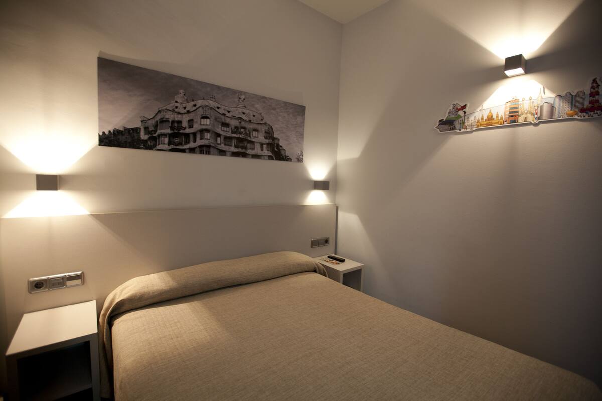 Quarto de hotel com cama, mesa de cabeceira, prateleira na parede com objetos decorativos e pintura de paisagem urbana.