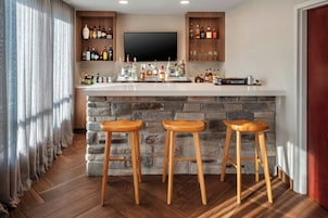 Uma área de bar com bancada de pedra, banquetas de madeira e prateleiras com garrafas de bebidas.