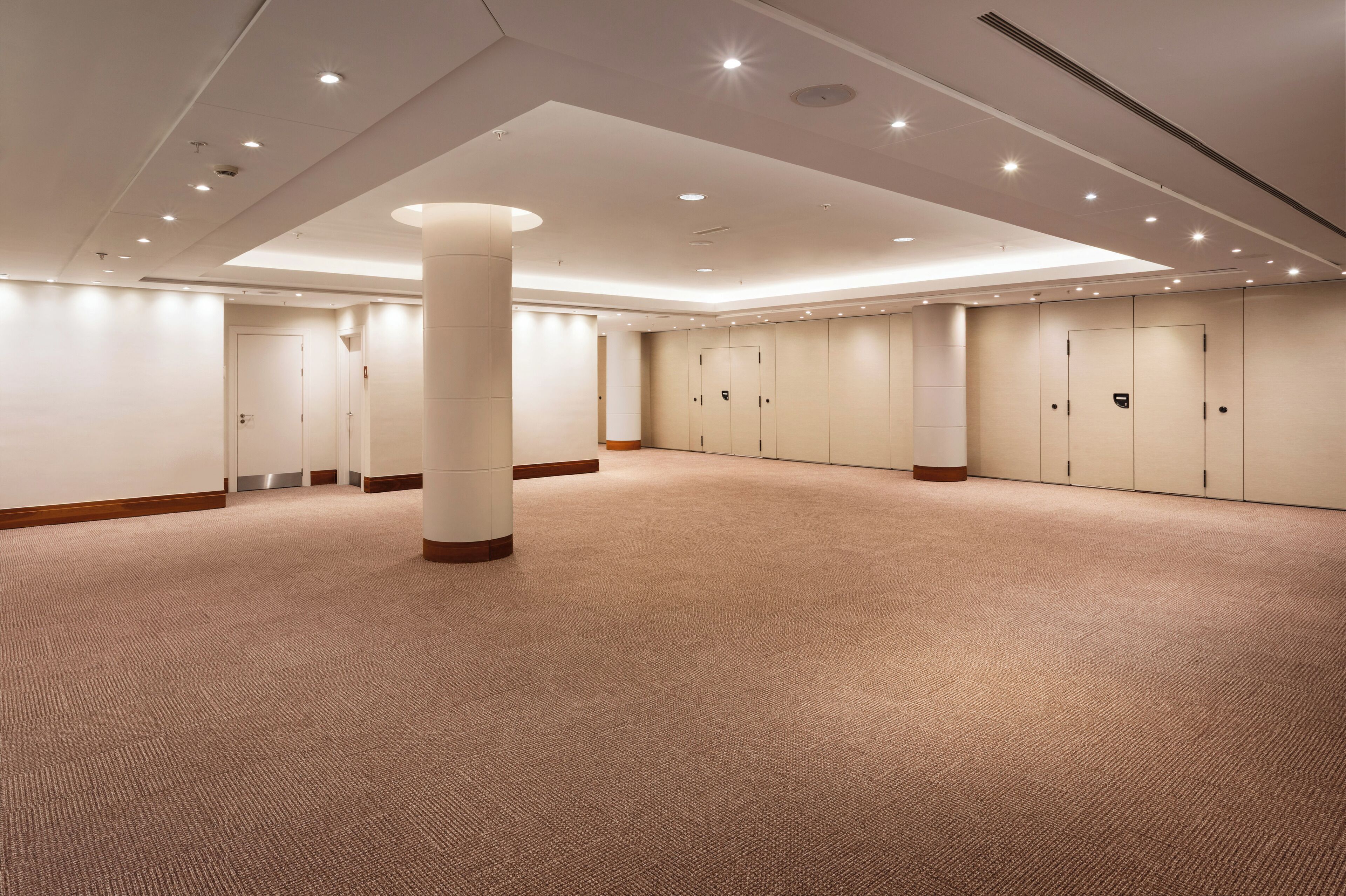 Uma sala de conferências ampla e bem iluminada, com piso coberto por carpete, várias portas e luminárias embutidas no teto.