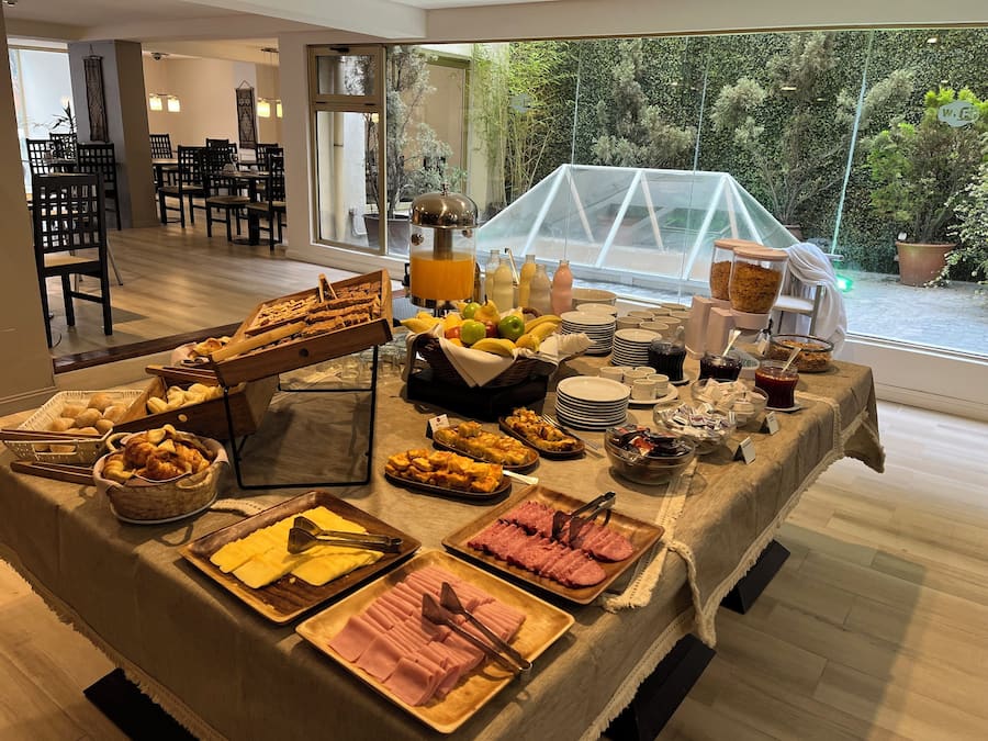 Uma mesa de buffet com diversos alimentos, incluindo doces, frutas e carnes, montada em um salão com grandes janelas que dão para um jardim.