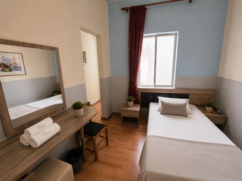 Quarto de hotel com cama, escrivaninha com espelho, janela com cortinas e uma pequena planta.