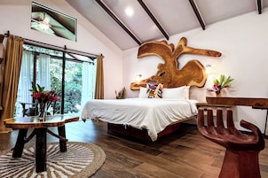Quarto com uma cama grande, cabeceira de madeira, uma mesa pequena com um vaso de flores e uma grande escultura de madeira.