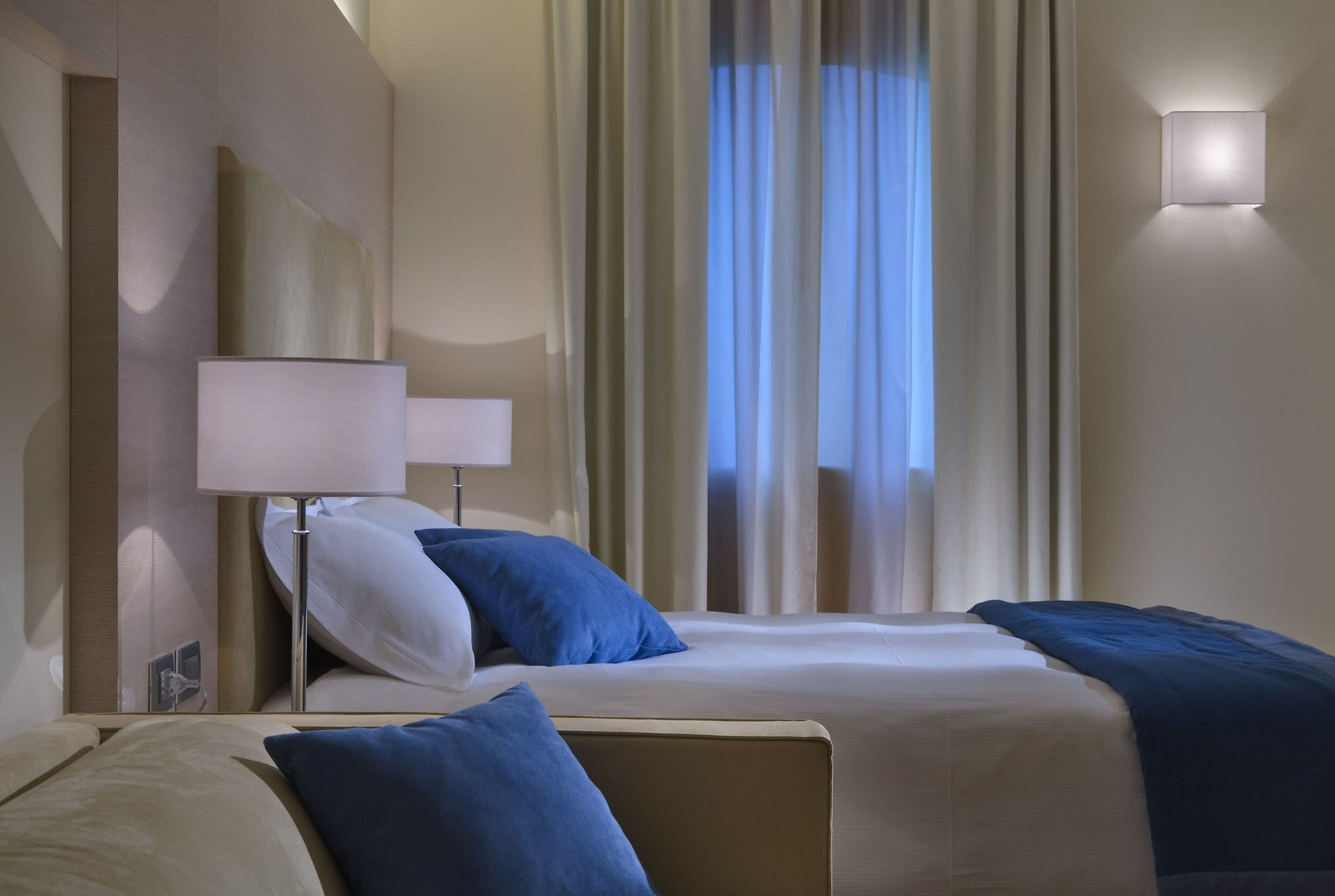 Quarto de hotel com duas camas, uma mesa de cabeceira com abajur e luminária na parede.