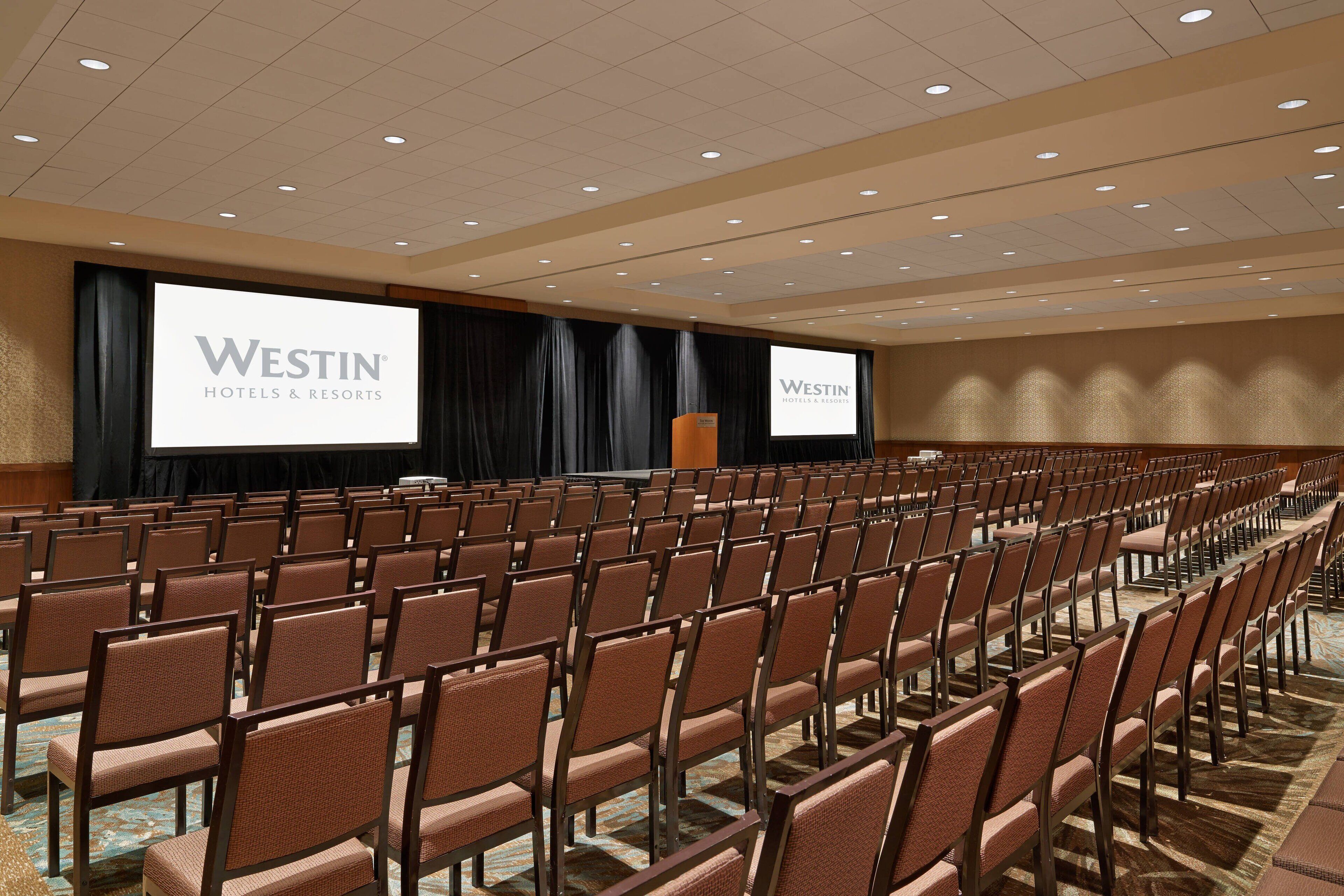 Uma sala de conferências com fileiras de cadeiras, uma tela grande exibindo o logotipo Westin Hotels & Resorts e um pódio.