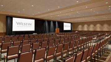 Uma sala de conferências com fileiras de cadeiras, uma tela grande exibindo o logotipo Westin Hotels & Resorts e um pódio.