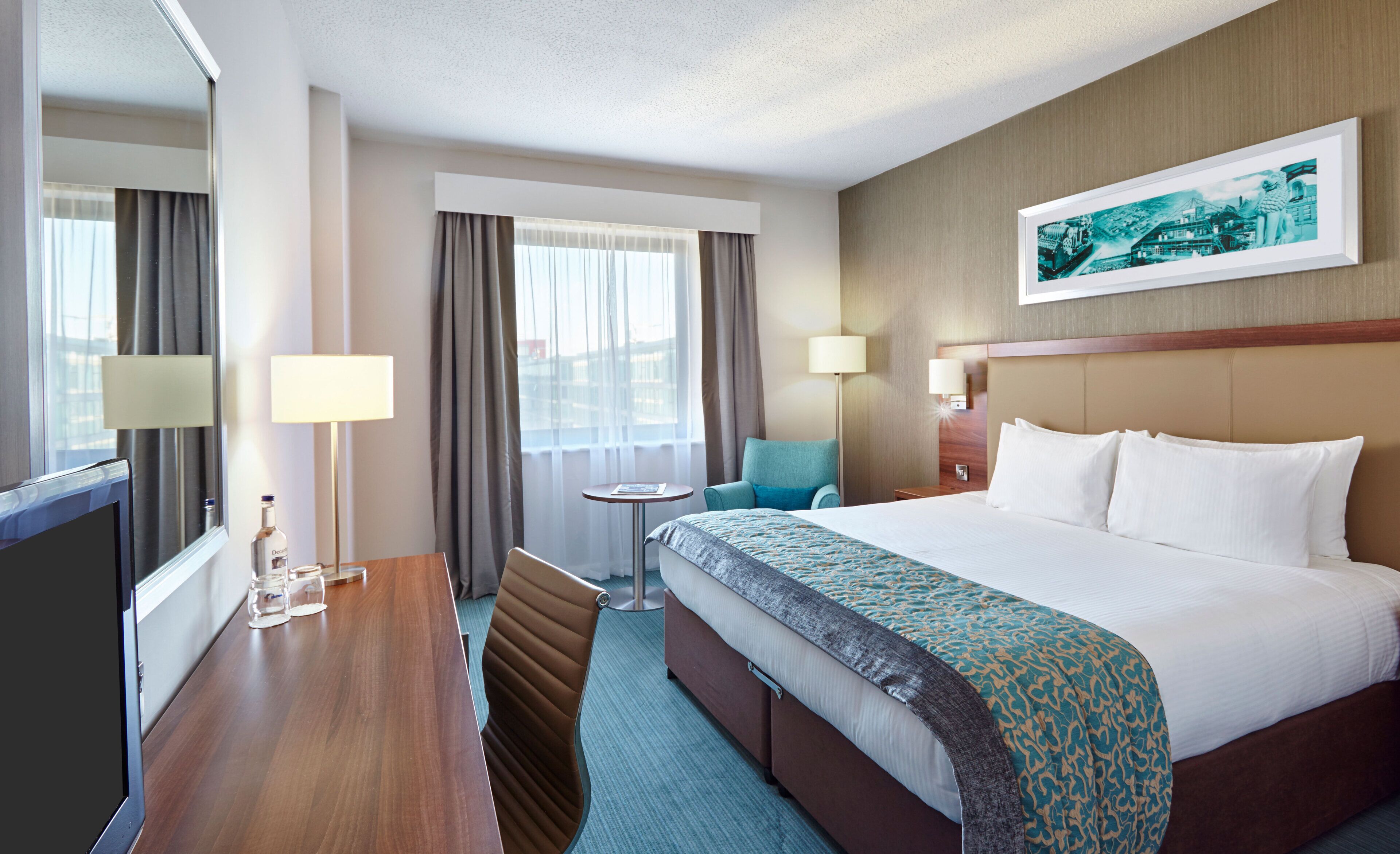 Quarto de hotel com cama, escrivaninha, cadeira, mesinha e janela com cortinas.