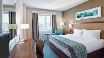 Quarto de hotel com cama, escrivaninha, cadeira, mesinha e janela com cortinas.
