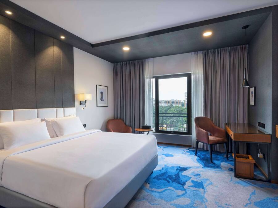 Quarto de hotel com uma cama grande, uma escrivaninha, uma cadeira, uma televisão e uma janela com cortinas.