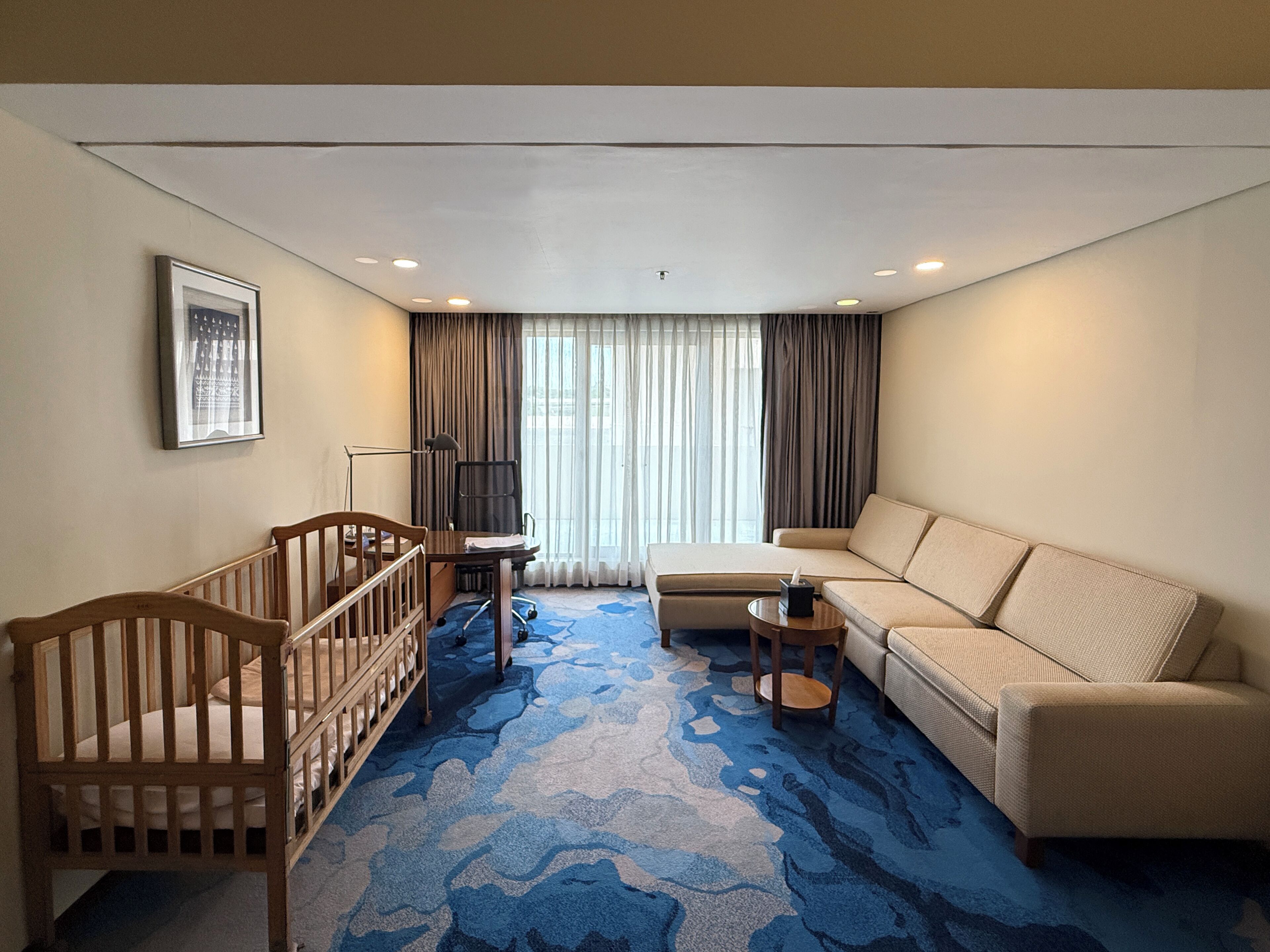 Quarto de hotel com berço, sofá, mesa pequena e abajur.