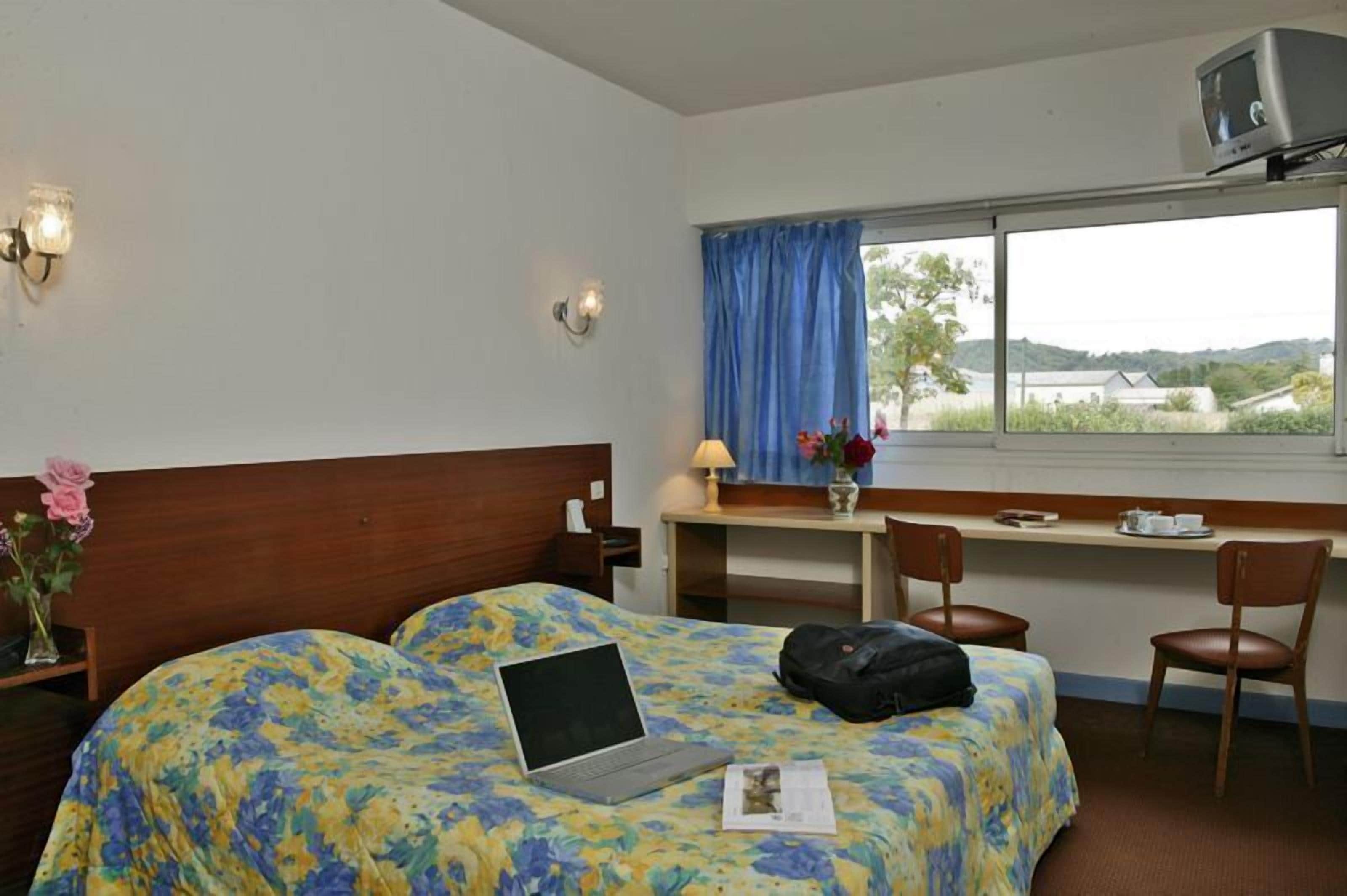 Quarto de hotel com cama, escrivaninha, laptop, cadeira, luminária e televisão.