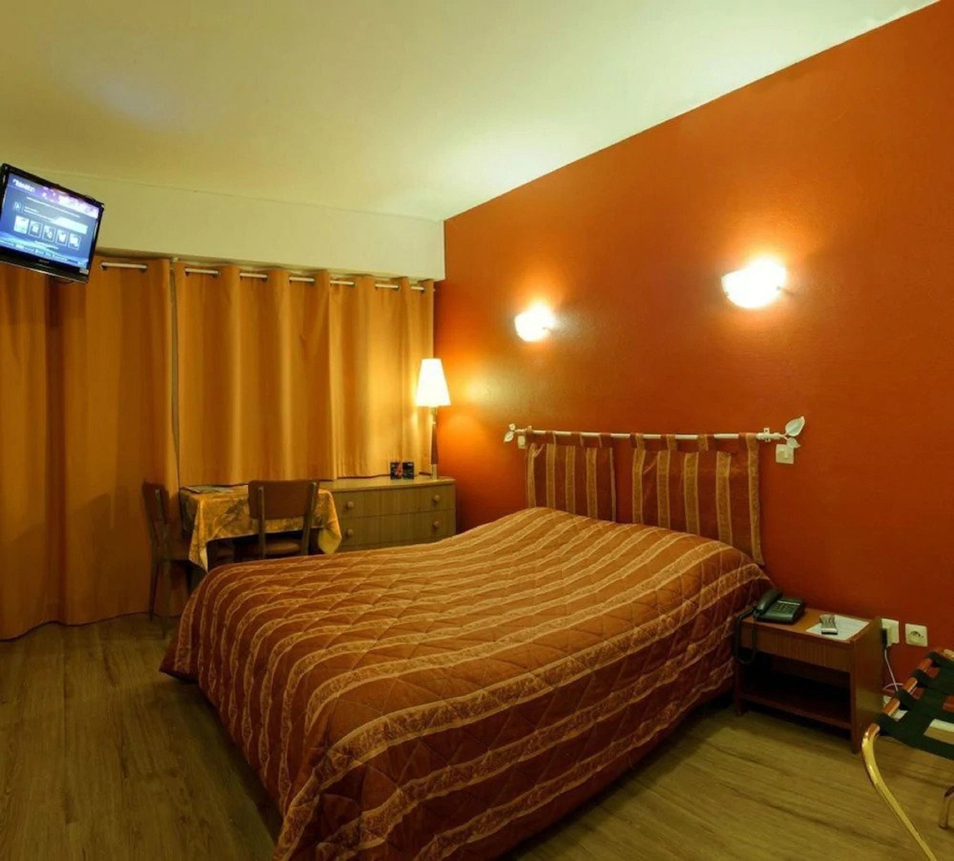 Quarto de hotel com cama, escrivaninha, cadeira, televisão e luminária.