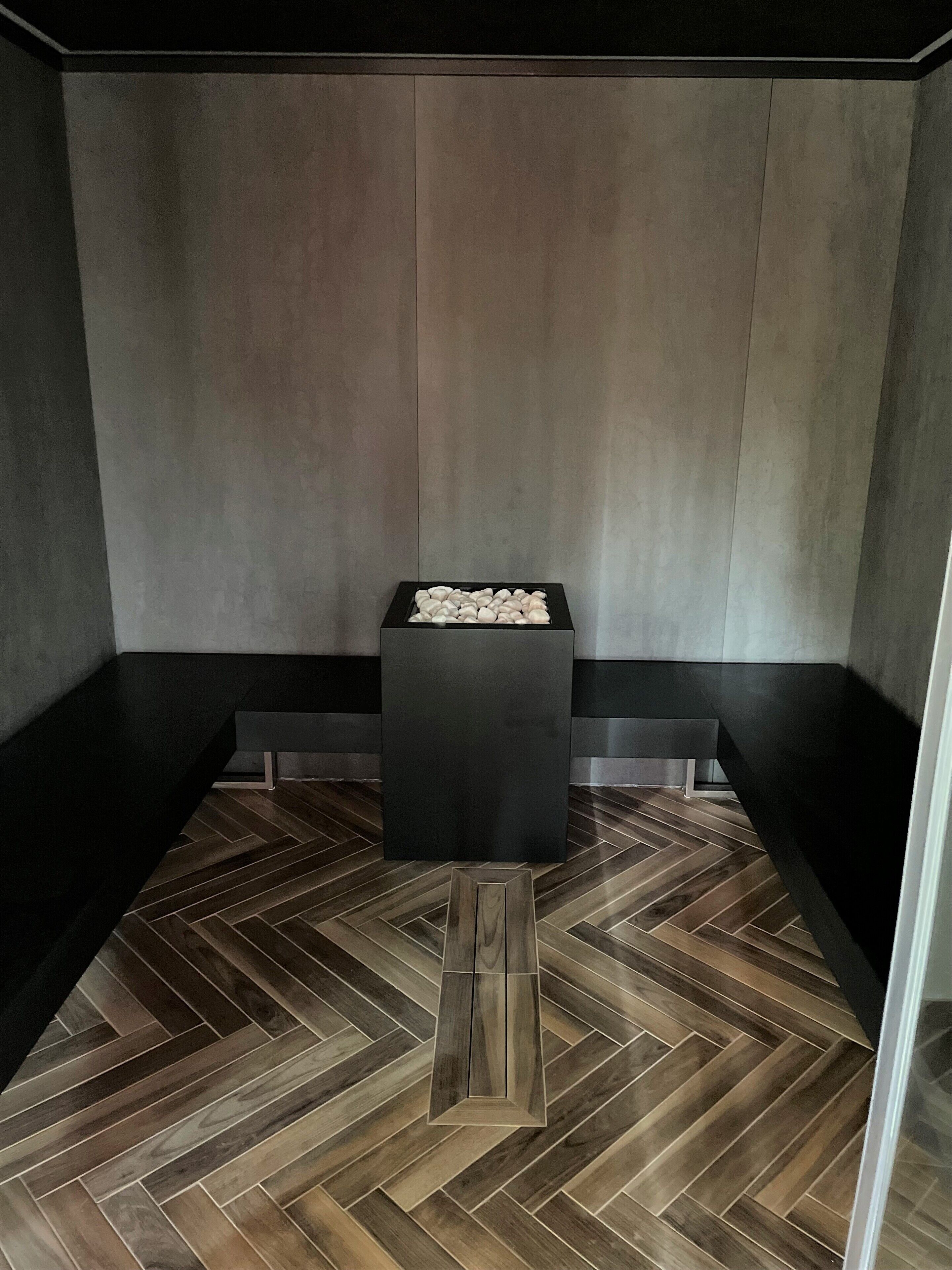 Quarto moderno e minimalista com piso de madeira em padrão herringbone, um banco preto e uma pequena mesa com um objeto branco.
