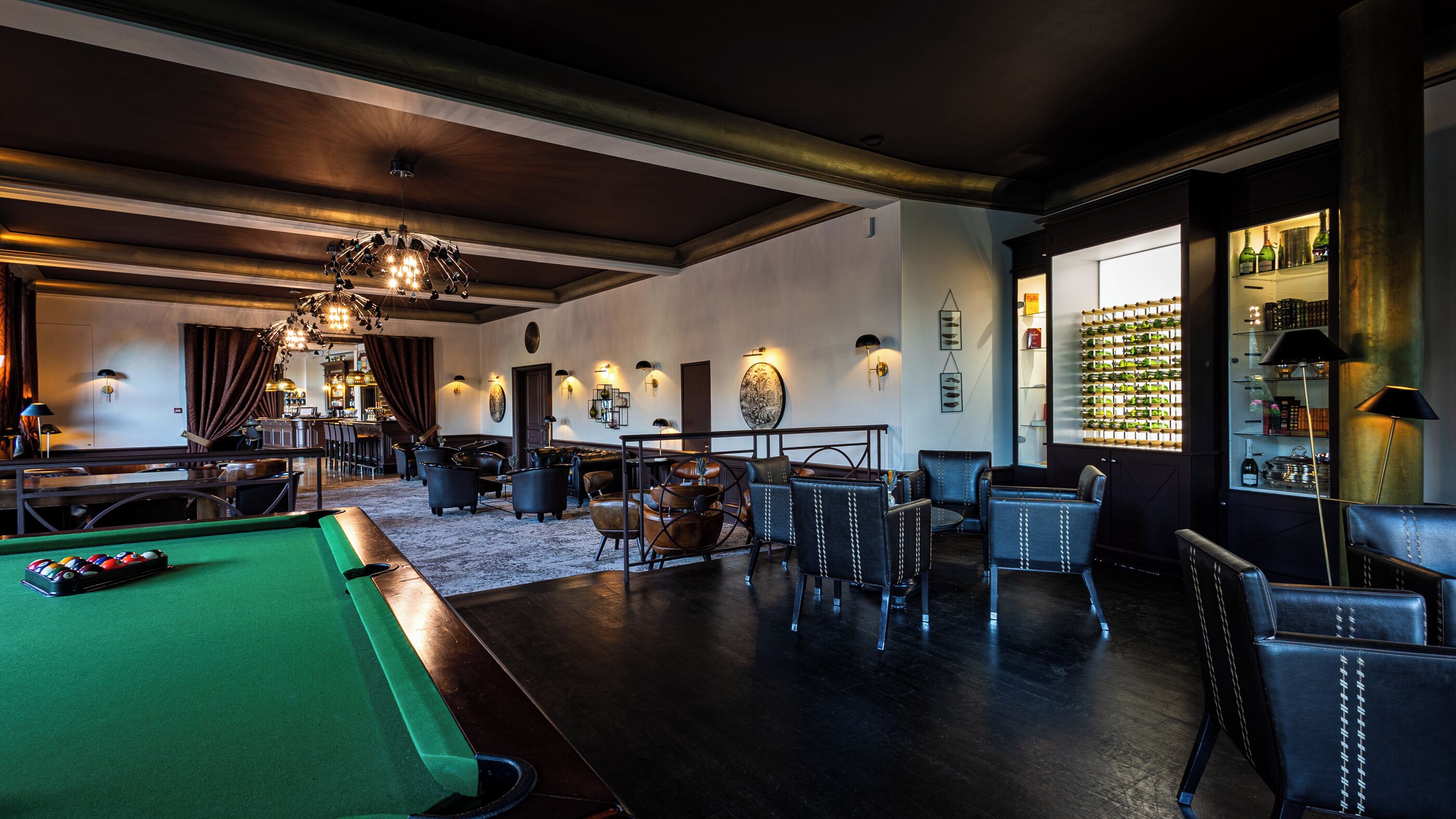 Uma sala de bilhar com mesa de sinuca, poltronas de couro e um espaço bar com garrafas.