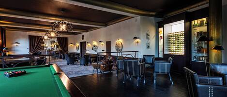 Uma sala de bilhar com mesa de sinuca, poltronas de couro e um espaço bar com garrafas.