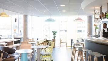 Zona de bar moderna com mesas redondas, banquetas e amplas janelas com vista para a cidade.