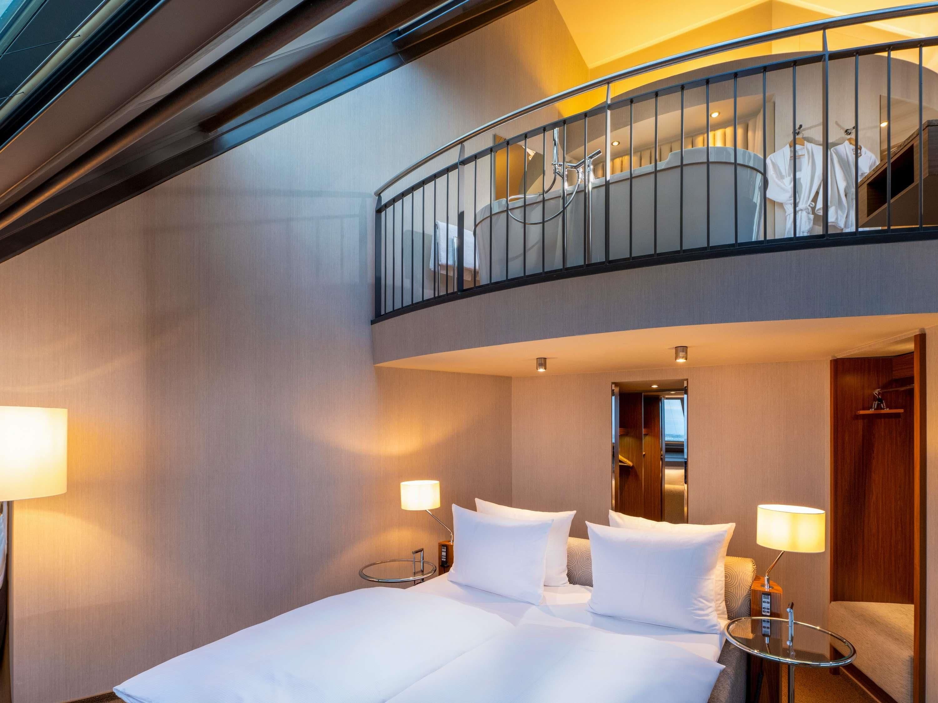 Quarto de hotel com uma cama grande, duas luminárias de cabeceira, uma mesa redonda e uma varanda com guarda-corpo.