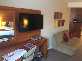 Quarto de hotel com televisão de tela plana na parede, uma escrivaninha de madeira com projetor, um sofá bege com almofadas vermelhas e um armário de madeira.