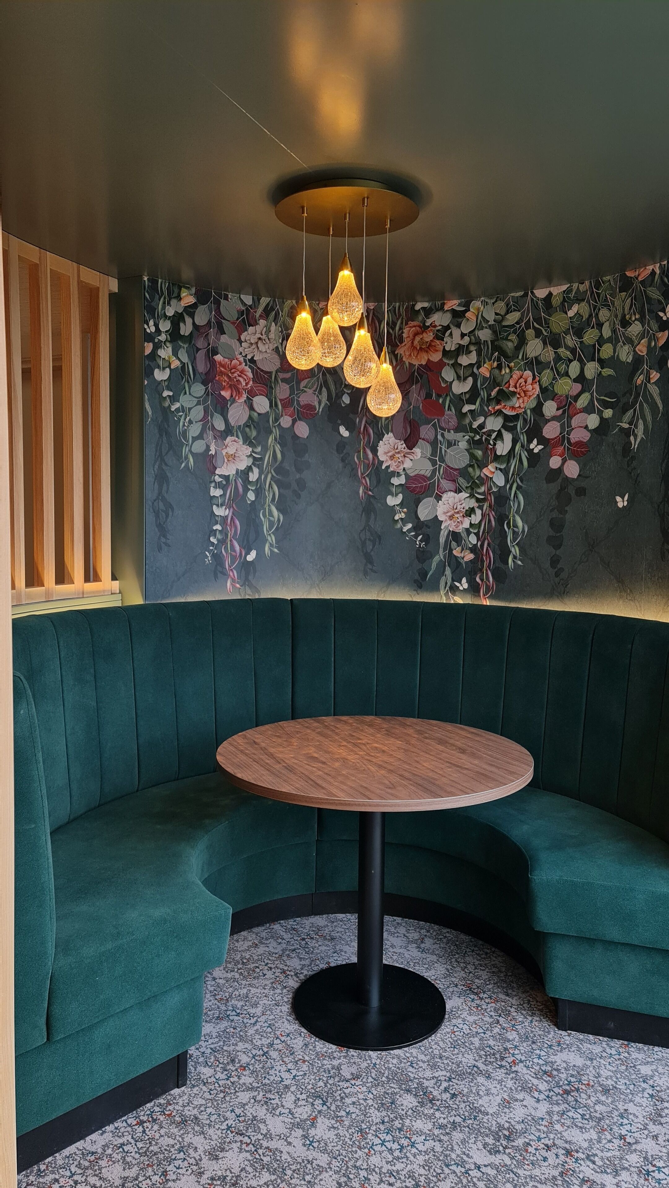 Um banco estofado verde com uma mesa redonda de madeira, um mural floral na parede e luminárias pendentes.